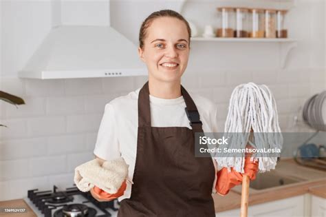 흰색 티셔츠와 갈색 앞치마를 입고 검은 머리를 한 웃는 여성의 실내 샷 손에 걸레와 천을 들고 가정부는 행복한 표정으로 카메라를