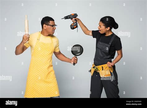 Angry African American Man In Apron Holding Rolling Pin And Frying Pan While Fighting With