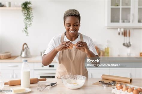 앞치마의 쾌활한 밀레니엄 세대 아프리카 계 미국인 여성이 미니멀리스트 주방 인테리어에서 반죽을 준비하는 달걀을 깨뜨립니다 25 29세에 대한 스톡 사진 및 기타 이미지