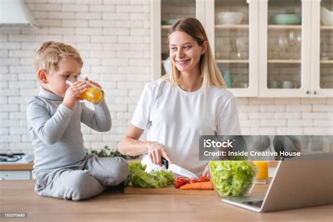 부엌에서 오렌지 주스를 마시는 쾌활한 작은 아이 아들은 함께 부엌에서 야채 샐러드를 요리하는 어머니를 돕습니다 가족 시간 건강한 식생활 개념 어버이날에 대한 스톡 사진 및