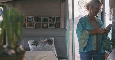 성인 여성은 미소를 지으며 휴대 전화를 사용하여 대체 집 캠핑카 밴의 문에 서서 메시지를 보냅니다 캐러밴으로 여행을 즐기고 로밍 연결을 사용하는 관광객 사진