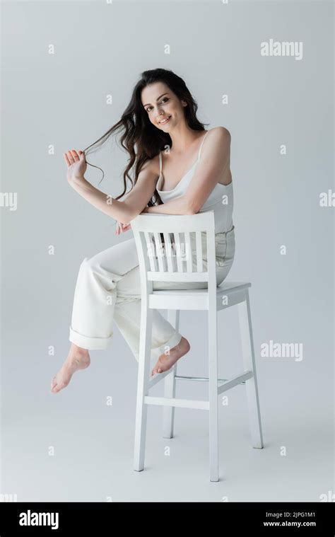 Full Length Of Positive Barefoot Woman Sitting On Chair On Grey