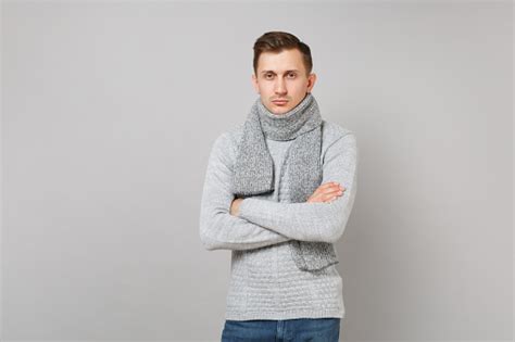 회색 스웨터에 심각한 젊은 남자 스튜디오에서 회색 벽 배경에 고립 된 손으로 접는 스카프를 들고 건강 한 패션 라이프 스타일