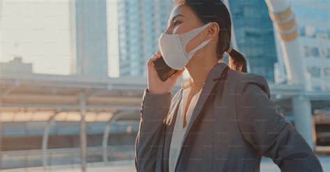 패션 사무실의 젊은 아시아 사업가가 도시 도시에서 혼자 야외를 걷는 동안 전화를 통해 의료 얼굴 마스크 이야기를 착용합니다