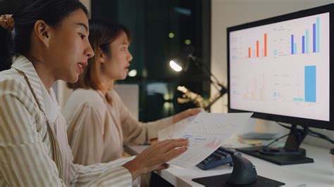 젊은 아시아 사업가들은 컴퓨터 모니터 앞에 앉아 프레젠테이션에서 차트 보고를 논의하는 화면을 보고 야간 근무 초과 근무 데이터를 비교합니다 늦게까지 일합니다 2명에 대한