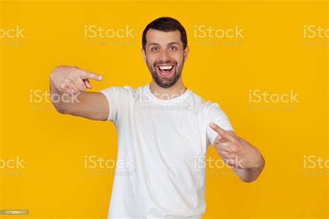 흰색 티셔츠에 수염을 기고 있는 청년이 행복하고 재미있는 얼굴로 카메라를 손가락으로 가리켰다 좋은 에너지와 분위기 격리된 노란색 배경에 서 T 셔츠에 대한 스톡 사진 및
