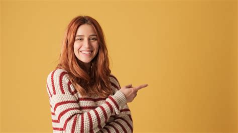 스웨터를 입은 빨간 머리 소녀는 스튜디오의 노란색 배경에 모형 공간을 보여주기 위해 시선을 돌렸습니다 아름 다운 소녀는 당신의 승진을 위해 노란 벽에 빈 공간이있는