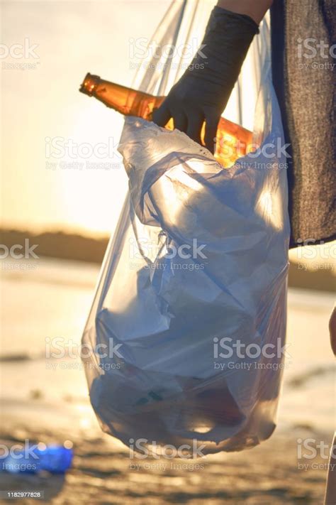 검은 장갑을 낀 젊은 자원봉사자가 강의 더러운 해변을 따라 쓰레기 봉투를 들고 걷고 쓰레기를 청소하고 있습니다 사람과 생태 클로즈업 가느다란 관에 대한 스톡 사진 및 기타