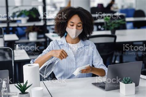 코로나바이러스로부터 건강을 보호하기 위한 직장 위생 마스크를 쓴 아프리카계 미국인 여성은 사무실 내부에서 책상과 노트북을 소독하기 위해 스프레이와 종이 타월을 사용합니다