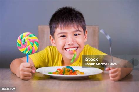 젊은 행복 하 고 흥분 남자 아이 명랑 먹는 접시 가득 사탕과 막대 사탕 설탕 중독 및 남용과 달콤한 건강에 해로운 영양에 회색 배경에 고립 된 테이블에 앉아 웃 고 먹기에