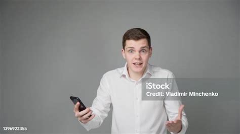 손에 휴대 전화를 들고 놀란 젊은 남자 그는 스마트 폰 화면에서 본 것에 놀랐습니다 회색 배경에 흰 셔츠를 입은 남자가 한 손에