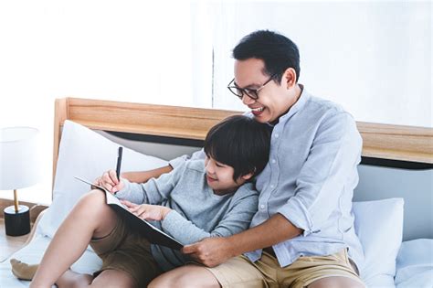 아버지와 아들은 함께 책에 숙제를 쓰고 침대에 앉아 아버지는 미소로 쓰는 아들을 가르치고있다 2명에 대한 스톡 사진 및 기타 이미지 Istock