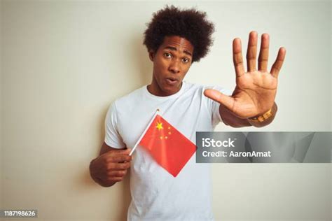 중국 국기를 들고 있는 젊은 아프로 미국인 남자가 고립 된 흰색 배경 위에 서 있고 진지하고 자신감있는 표정으로 정지 표지판을하고 방어 제스처 경계 표지에 대한 스톡 사진