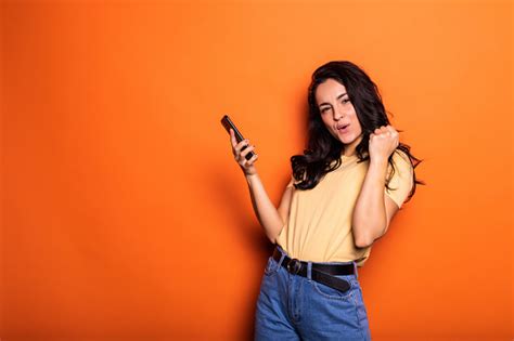 스마트 폰 메시지를 읽고 예 제스처를 보여주는 젊은 여자 좋은 소식 오렌지 배경에 고립 된 내기 응용 프로그램 사람들에 대한 스톡 사진 및 기타 이미지 Istock