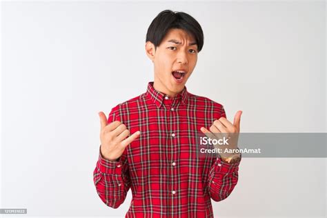 고립 된 흰색 배경 위에 서있는 캐주얼 한 빨간 셔츠를 입은 젊은 중국 남자가 미친 표정으로 손을 들고 바위 기호를하고 있습니다 음악 스타 무거운 개념 개념에 대한 스톡