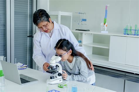 과학 교실에서 학생들의 학습 안경을 쓴 집중된 소녀는 노트북과 실험실 장비가 있는 교실에서 흰색 실험복을 입은 아시아 교사의 안내를 받아 현미경을 통해 검사합니다 55 59