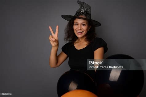 할로윈 파티를위한 마녀 카니발 의상을 입은 마법사 모자를 쓴 이빨이 있는 미소 여자와 함께 아름다운 미소가 있는 아름다운 미소는 평화 표지판을 보여주고 검은 풍선으로 회색