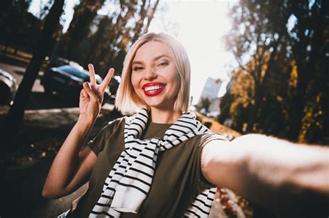 예쁜 젊은 금발 여자가 셀카를 찍고 사진을 찍고 V 사인을 입고 카키색 옷을 입고 어깨를 묶고 스웨터를 걷고 도시 도시 공원 밖으로 걷는 사진 20 24세에 대한 스톡 사진