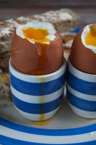 파란색과 흰색 코니쉬 도자기 그릇 차 접시 달걀 컵 부드러운 삶은 달걀 통밀 버터 구운 빵 조각 콧물 노란색 노른자 나무 곡물