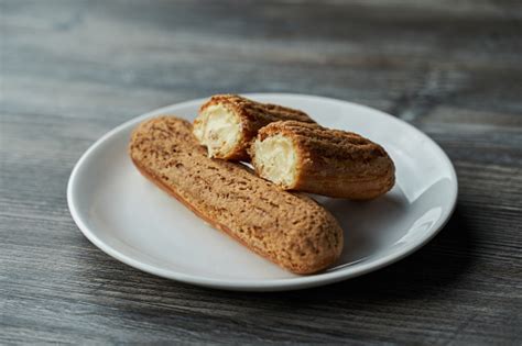 흰색 접시에 커스터드가 있는 두 개의 에클레어 하나는 반으로 자르고 두 번째에 놓여 있습니다 회색 나무 테이블에 접시 스탠드 가루 설탕에 대한 스톡 사진 및 기타 이미지