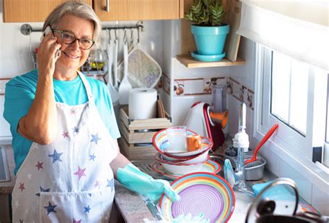 한 선임 여성이 전화를 받기 위해 판세척기를 멈춘다 회색 머리와 안경 앞치마와 장갑을 착용하여 설거지 부엌의 코너입니다 직장에서 할머니 창에서 밝은 빛 가사 집안일에 대한