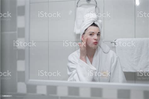 흰색 목욕 가운을 입고 머리에 수건을 두른 젊은 백인 여성이 얼굴에서 보습 시트 마스크를 제거하고 욕실에서 뷰티 트리트먼트를