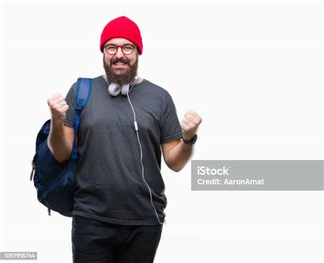 축 하 하는 고립 된 배경 위에 빨간 모직 모자와 배낭을 입고 젊은 Hipster 남자 놀라게 하 고 팔 제기 성공을 위한 놀라게 했 고 눈 우승자 개념입니다 과체중에 대한