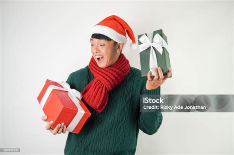 산타 클로스 모자 빨간 스카프 녹색 긴팔 스웨터를 입고 웃는 얼굴 표정을 가진 젊은 아시아 남자가 흰색 배경에 고립 된 리본이 달린 큰 선물 상자를 들고 있습니다 12월에