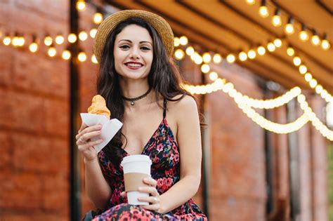 어두운 눈을 가진 젊은 여성 모델 여름 드레스와 모자를 입고 빨간 입술을 입고 손 테이크 아웃 코프와 크루아상을 들고 야외 레스토랑에서 아침 식사를합니다 음식과 휴식 개념