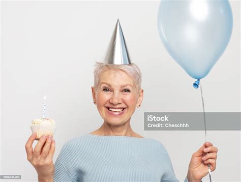 촛불과 파란색 풍선이 달린 컵케이크로 생일을 축하하는 원뿔형 모자를 쓰고 행복한 노인 여성 유머에 대한 스톡 사진 및 기타