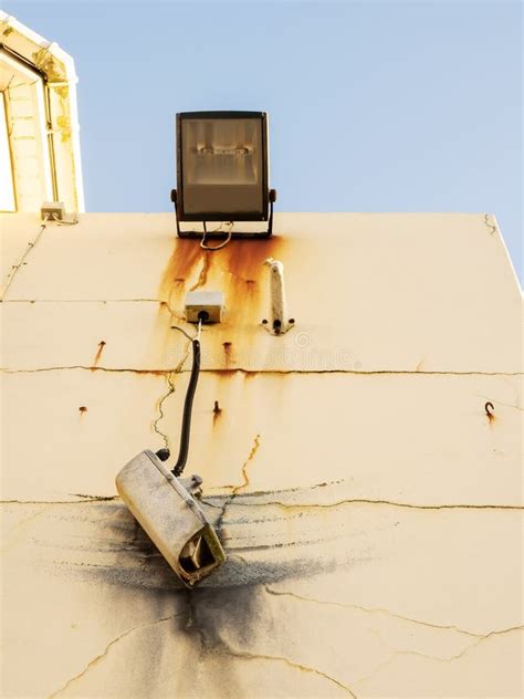 Damaged CCTV Camera Hanging On A Cable On Yellow Wall Old Flood Light Above The Camera Front