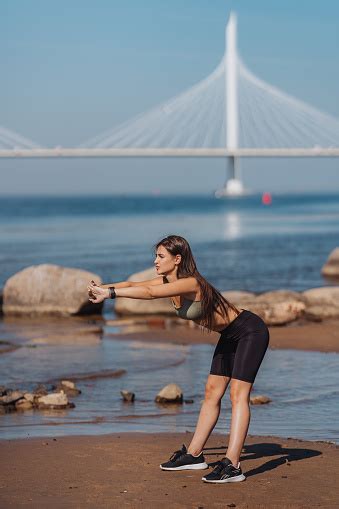 갈색 머리의 미국 적합 소녀는 운동복을 입고 화창한 여름날에 만 다리에 대한 해변에서 스트레칭 운동을 합니다 예쁜 백인 젊은 여자가 야외에서 워밍업하고 있습니다 피트니스
