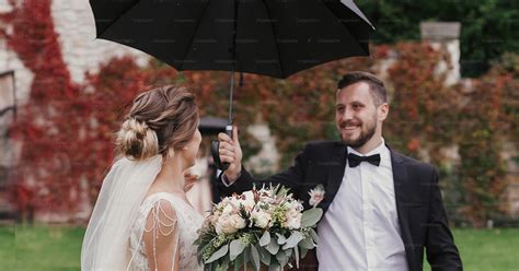 세련 된 신랑은 비오는 야외에서 우산 아래에서 화려한 신부와 미소를 짓고 있습니다 관능적인 결혼식 커플은 빗속에서 즐거운