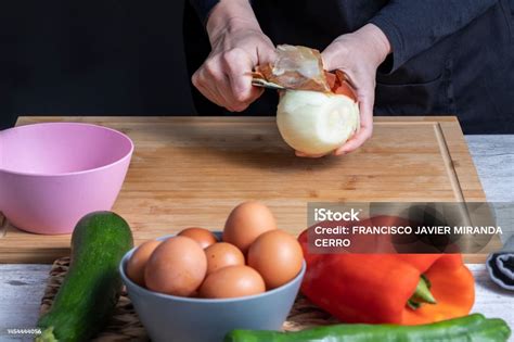 여자의 손은 칼을 사용하여 나무 부엌 보드에 양파를 껍질을 벗기고 있습니다 계란 빨간색과 녹색 피망 양파와 호박이 들어간 흐릿한 분홍색과 회색 그릇 가구에 대한 스톡 사진