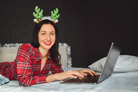 빨간 잠옷을 입은 아름다운 흥분된 웃는 갈색 머리 여자는 크리스마스 순록 뿔의 침대에 누워 어두운 실내 침실에서 노트북으로 작업합니다 새해 복 많이 받으세요 분위기에서
