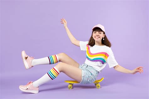 스튜디오에서 보라색 파스텔 벽 배경에 고립 된 손을 확산 노란 스케이트 보드에 앉아 생생한 옷에 재미 귀여운 십 대 소녀 사람들은 진심으로 감정 라이프 스타일 개념입니다