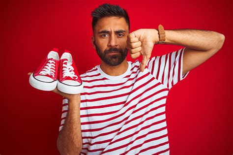 젊은 아랍 인도 히스패닉 남자 들고 스포츠 캐주얼 운동화 위에 고립 된 빨간 배경 화가 얼굴 엄지 손가락으로 싫어하는 표시