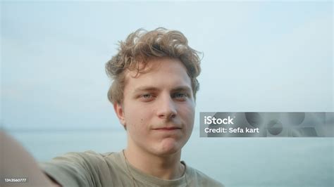 잘 생긴 젊은 금발 머리 색깔의 눈 남자 는 카메라에 손을 흔들고 미소 그의 손에 전화로 화상 통화 채팅을만드는 사람 바다 바다 배경 Brand Name Smart