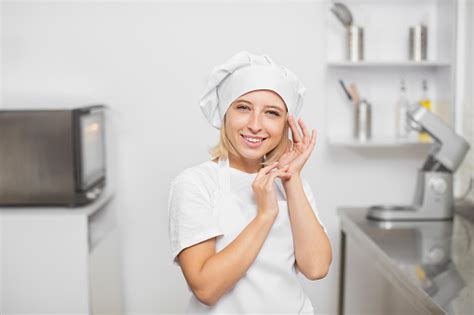 사람 직업 및 작업 개념 젊은 꽤 즐거운 여자 과자 흰색 앞치마와 모자를 입고 미소와 함께 카메라에 포즈 현대 부엌 인테리어에