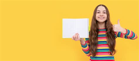 행복한 아이는 복사 공간 노란색 배경을위한 열린 책을 들고 손 제스처를 엄지 손가락으로 주며 만족합니다 여학생 학생의 수평 고립 포스터 여학생의 배너 헤더 초상화 복사 공간