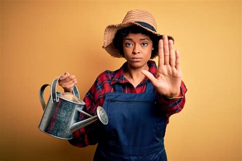 곱슬 머리를 가진 젊은 아프리카 계 미국인 농부 여자 앞치마와 모자를 사용 하 여 물 수 수 열려 있는 손으로 하 고 정지 기호를 심각 하 고 자신감 있는 표정 방어 제스처