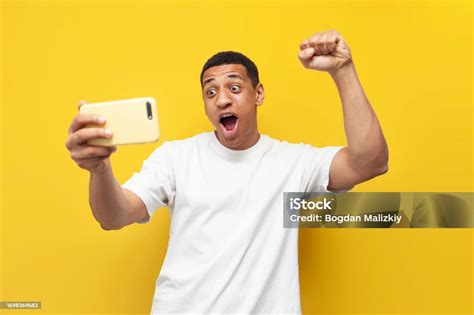 흰 티셔츠를 입은 젊은 아프리카계 미국인 남성이 스마트폰으로 모바일 게임을 하고 승리를 기뻐한다 비명에 대한 스톡 사진 및 기타