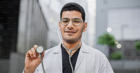 미소 짓는 친절한 남성 아랍 힌두교 의사 또는 의대생이 현대 병원 밖에 서서 청진기를 카메라에 시연하는 초상화 청진기에 대한 선택적 초점 사진 Unsplash의