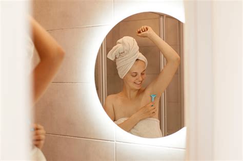 낙관적인 행복한 젊은 성인 여성은 욕실에서 겨드랑이를 면도하고 몸을 보고 탈필 과정을 즐기고 맨손으로 포즈를 취하고 흰색 수건으로 싸서 거울에 대한 스톡 사진 및 기타