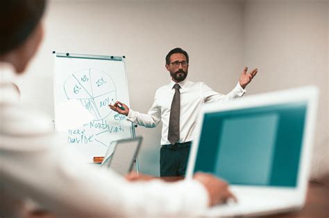 최고 는 동료들에게 사업 계획을 제시하고 있습니다 넥타이가 달린 흰색 셔츠를 입은 남자가 다이어그램을 들고 칠판에 서 있다 사무실 직원들은 회의에서 노트북이 있는 테이블에