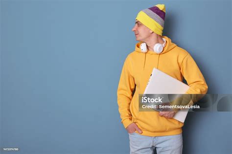 비니 모자와 캐주얼한 노란색 까마귀를 입은 귀여운 긍정적인 남자의 초상화는 파란색 배경 위에 고립되어 서 있고 복사 공간을 바라보고 노트북 광고 영역을 들고 있다 35 39