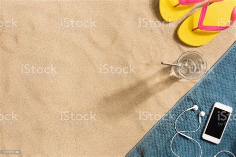 여름 방학 구성 플립 플롭 스마트 폰과 모래 배경에 물 한 잔 그림자가 있는 가혹한 빛 여름 배경입니다 수건으로 만든 국경