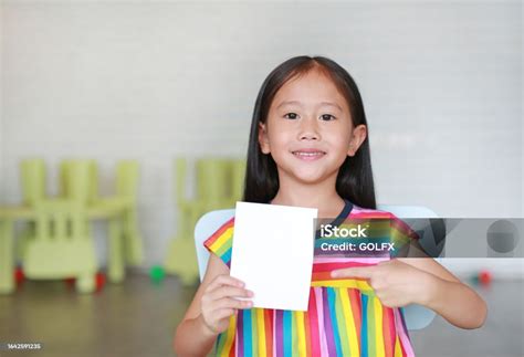 초상화는 웃고 있는 아시아의 어린 소녀가 빈 흰 종이 카드를 손에 들고 가리키고 있다 어린이 방에서 빈 종이 메모 복사 공간을 보여주는 아이 아이에 대한 스톡 사진 및 기타