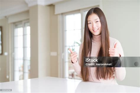 흰색 테이블에 캐주얼 스웨터를 입고 아름다운 아시아 여성은 손 엄지 손가락으로 긍정적 인 제스처를하고 성공 표지판미소와 행복 쾌활한 표정으로 카메라를 바라보며 승자 제스처