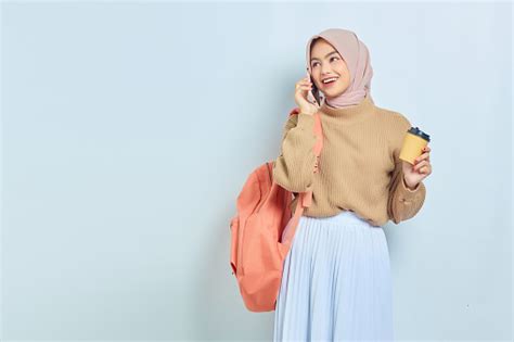 배낭을 들고 갈색 스웨터를 입은 젊은 아시아 무슬림 여성 학생을 웃고 휴대 전화로 이야기하고 흰색 배경에 고립 된 커피 나 차 한 잔을 들고 학교 개념으로 돌아가기 20